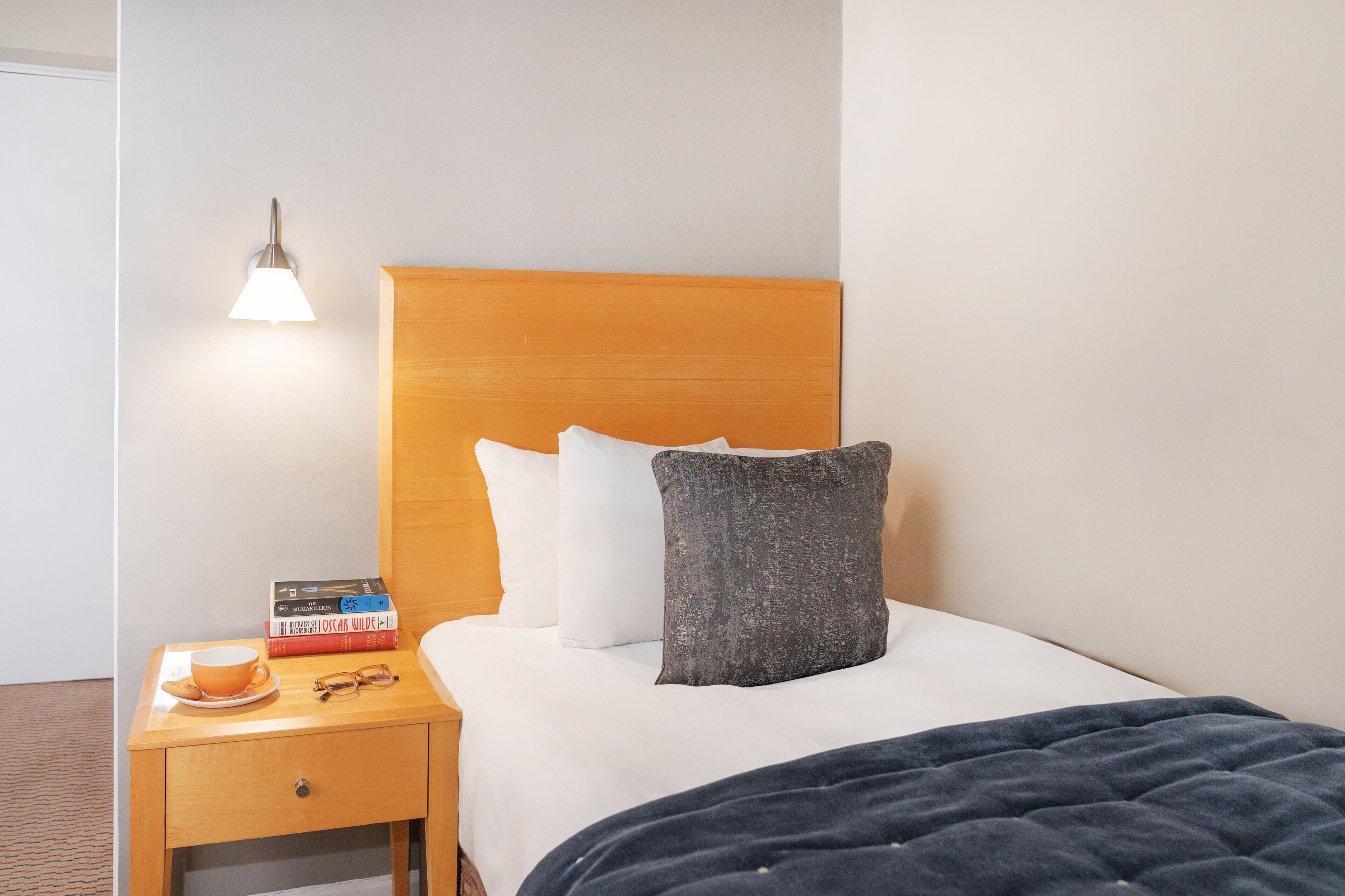 Cama bem arrumada com cabeceira de madeira, mesa de cabeceira com abajur e uma pilha de livros.