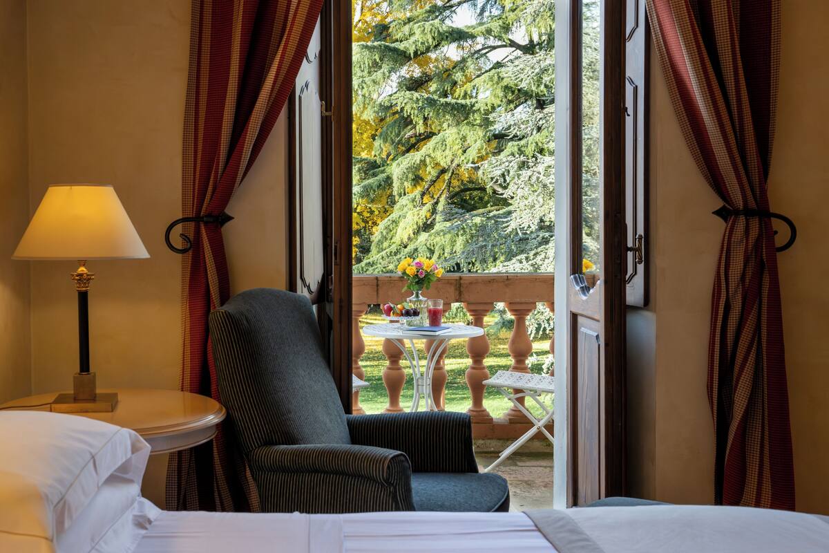 Quarto de hotel com cama, uma cadeira, uma mesa com um vaso de flores e vista para um jardim através de uma porta aberta.
