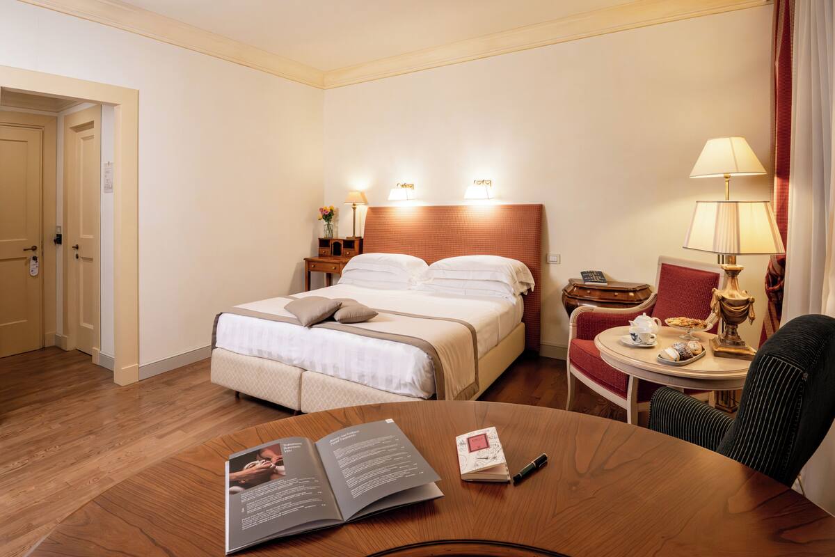 Quarto de hotel com cama, mesas de cabeceira, uma mesa redonda com uma cadeira e um livro sobre a mesa.