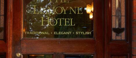 Entrada de madeira de um hotel com porta de vidro exibindo o nome "The Lyons Hotel" e um sinal de classificação de higiene alimentar.