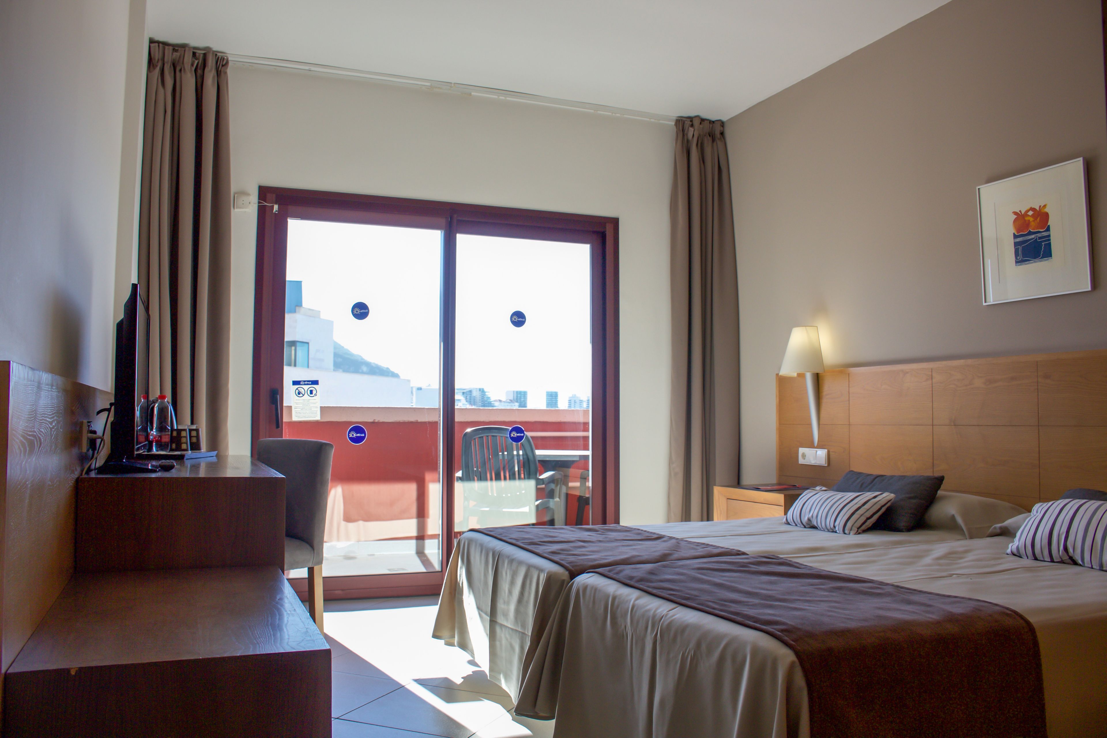 Quarto de hotel com cama, escrivaninha, cadeira, varanda e vista para edifícios, além de luminária na parede.