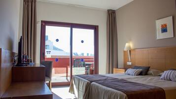 Quarto de hotel com cama, escrivaninha, cadeira, varanda e vista para edifícios, além de luminária na parede.