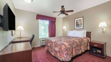 Quarto de hotel com cama, escrivaninha, cadeira, televisão, ventoinha no teto, janela com cortinas e mesa de cabeceira com abajur.