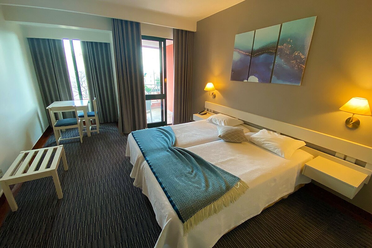 Quarto de hotel com duas camas, uma mesa e uma cadeira. Há duas luminárias de cabeceira e um quadro na parede.