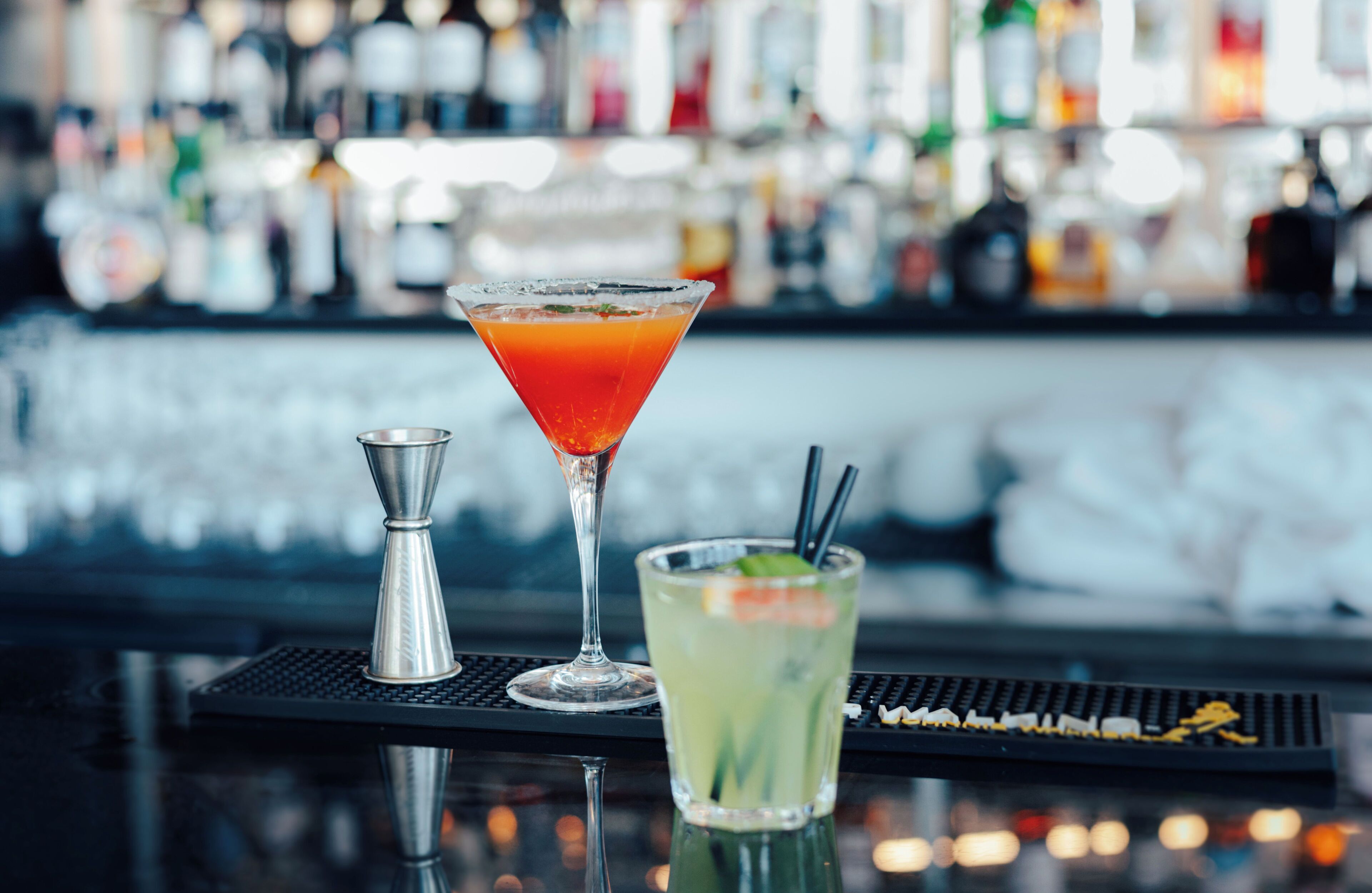 Ambiente de bar com uma taça de martini, um copo de cocktail e um coqueteleira sobre uma superfície refletora.