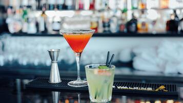 Ambiente de bar com uma taça de martini, um copo de cocktail e um coqueteleira sobre uma superfície refletora.