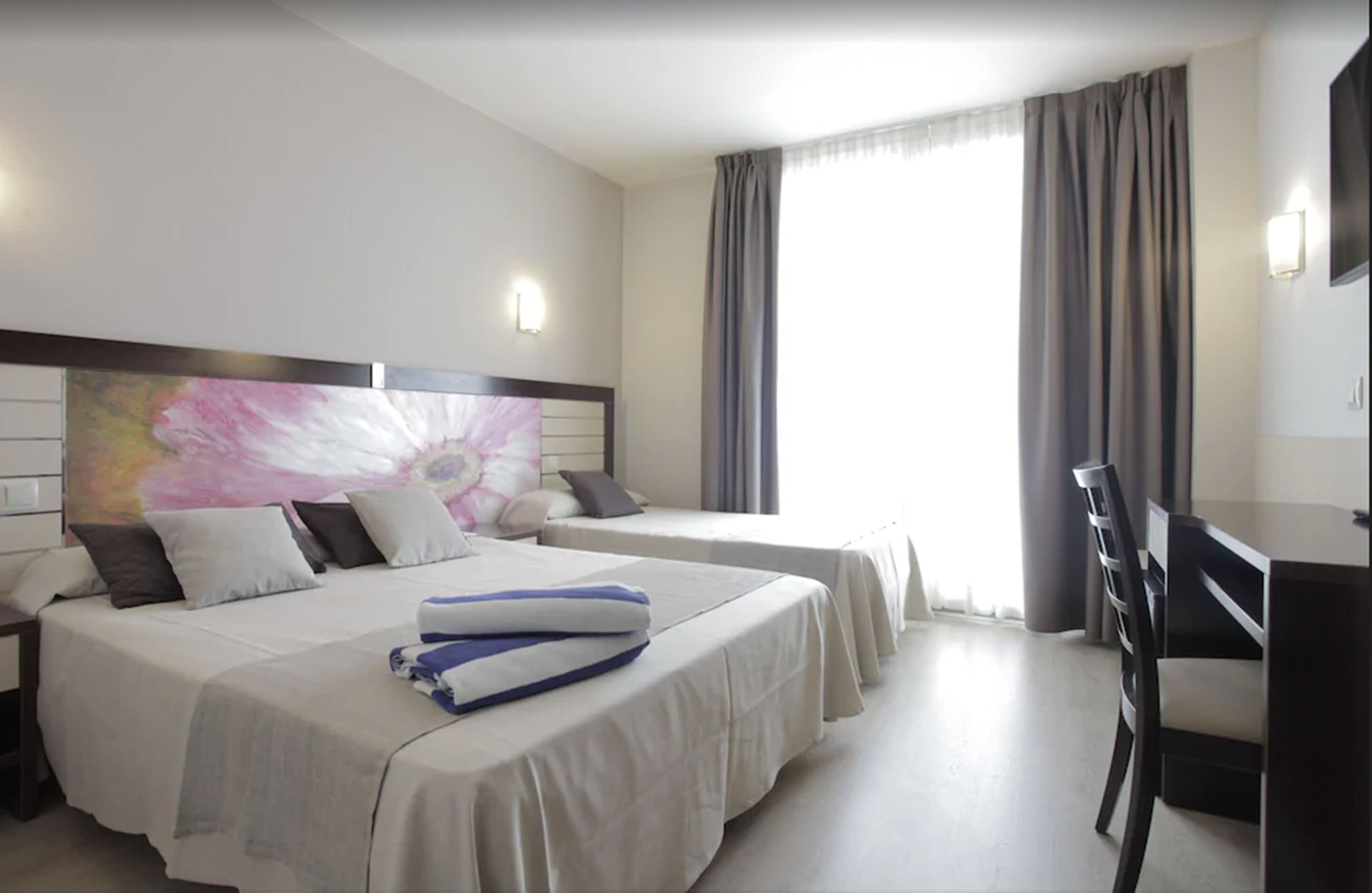 Quarto de hotel com duas camas, uma escrivaninha e uma janela grande com cortinas.