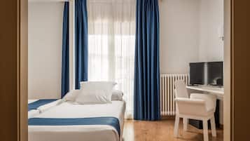 Quarto de hotel com uma cama, uma escrivaninha com cadeira, televisão e uma janela com cortinas.
