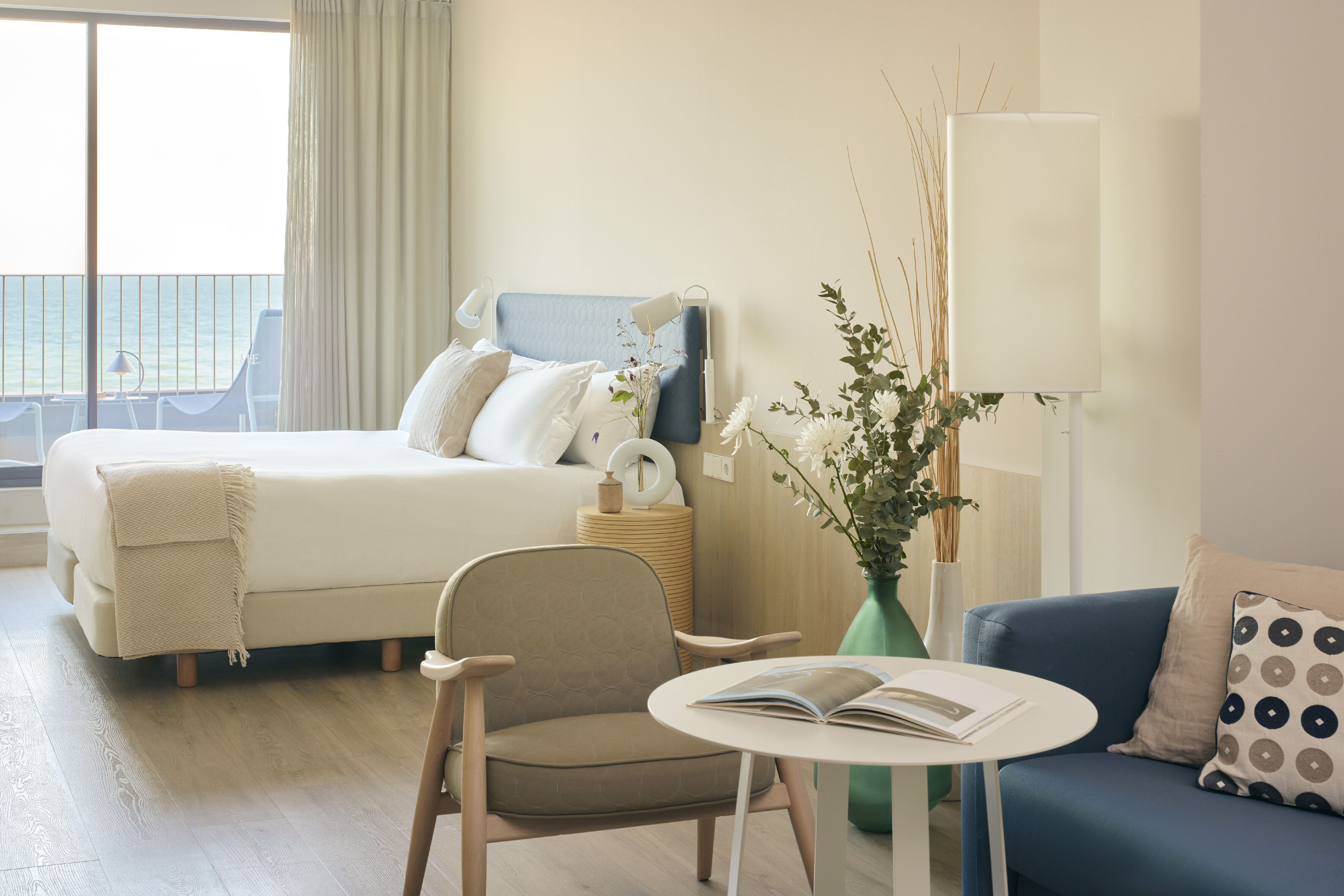 Quarto de hotel com cama, uma cadeira, uma mesinha com um livro, abajur e vista para o mar.