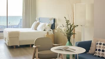 Quarto de hotel com cama, uma cadeira, uma mesinha com um livro, abajur e vista para o mar.