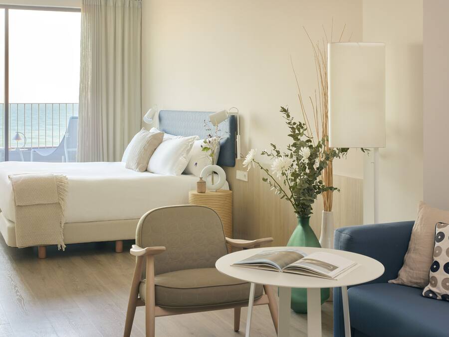 Quarto de hotel com cama, uma cadeira, uma mesinha com um livro, abajur e vista para o mar.