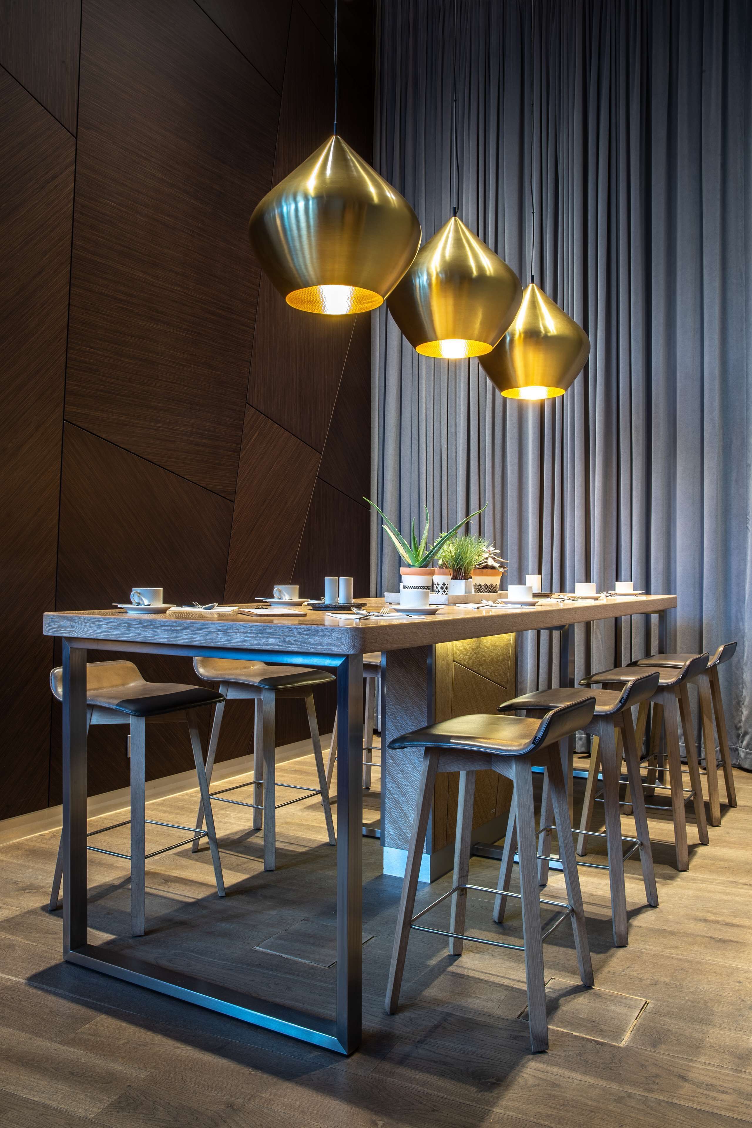 Uma sala de jantar moderna com uma mesa de madeira comprida, cadeiras de metal e três luminárias pendentes douradas.
