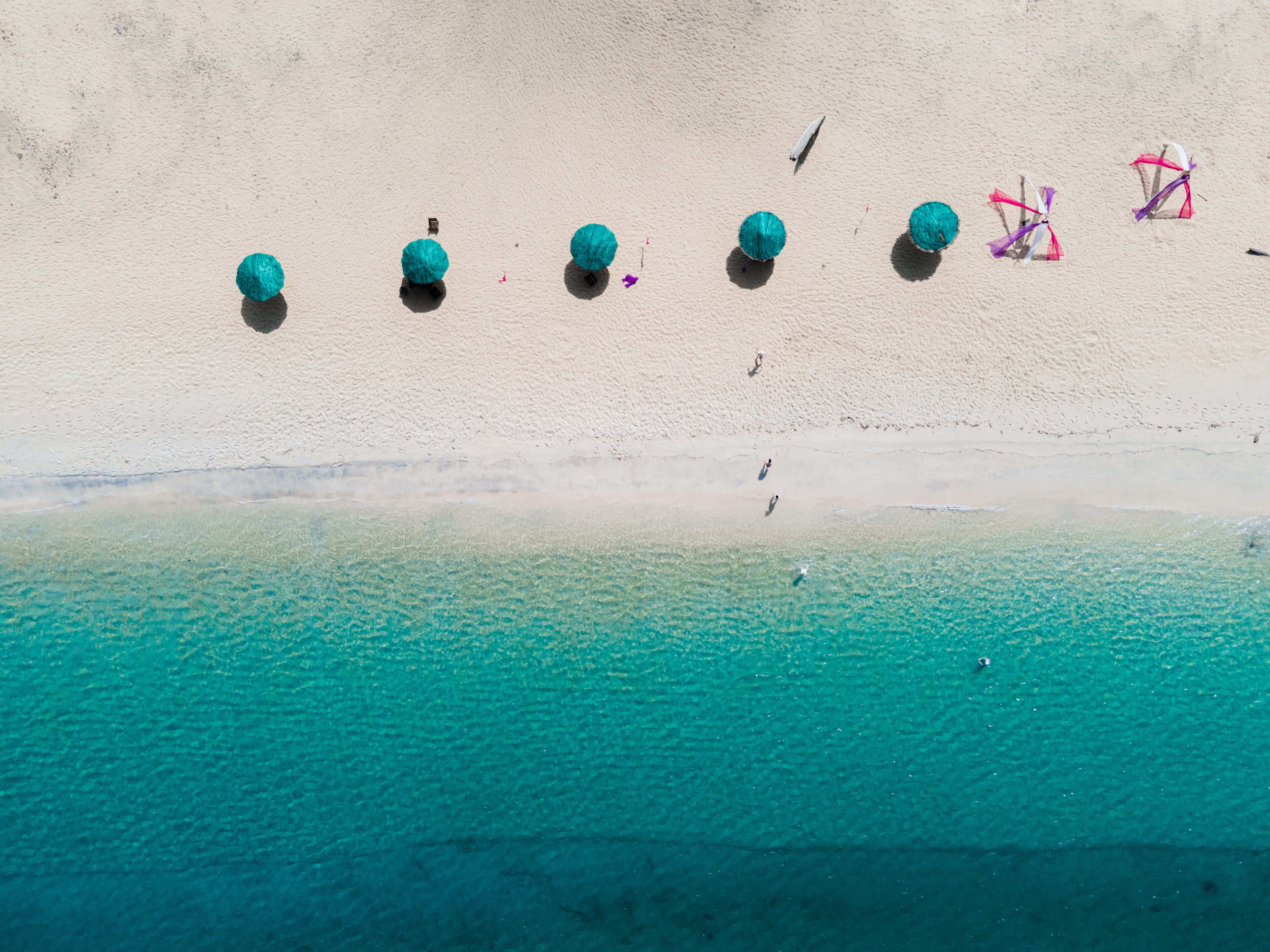 Vista aérea de uma praia com águas turquesa cristalinas e areia branca. Vários guarda-sóis coloridos e algumas pessoas podem ser vistas na areia.