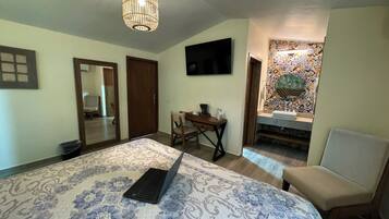 Quarto com cama, escrivaninha, cadeira, televisão e banheiro com painel de azulejos.