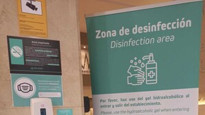 Entrada de hotel com um grande letreiro verde indicando uma área de desinfecção e instruções para usar álcool em gel. Há também avisos lembrando os hóspedes de usar máscaras e manter distância segura.