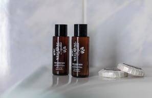 Duas embalagens de gel de banho e corpo 'Welcome' e duas barras de sabonete brancas sobre uma superfície refletora.