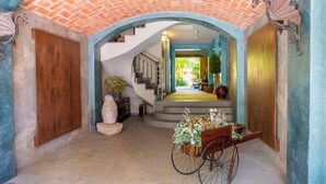 Um corredor com arco de tijolos, porta de madeira, escultura de pedra, carrinho de bicicleta com flores e escadaria que leva a um quarto iluminado com plantas.