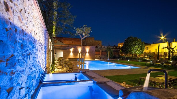 Área de piscina ao ar livre à noite, com muro de pedra, fonte de água e espreguiçadeiras.