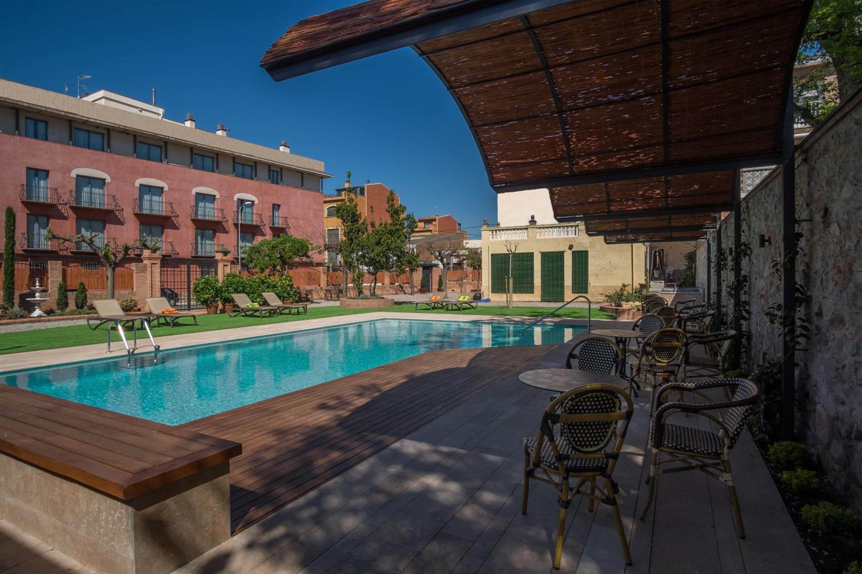 Piscina com espreguiçadeiras e mesas, rodeada por edifícios com varandas e um espaço coberto com alpendre.