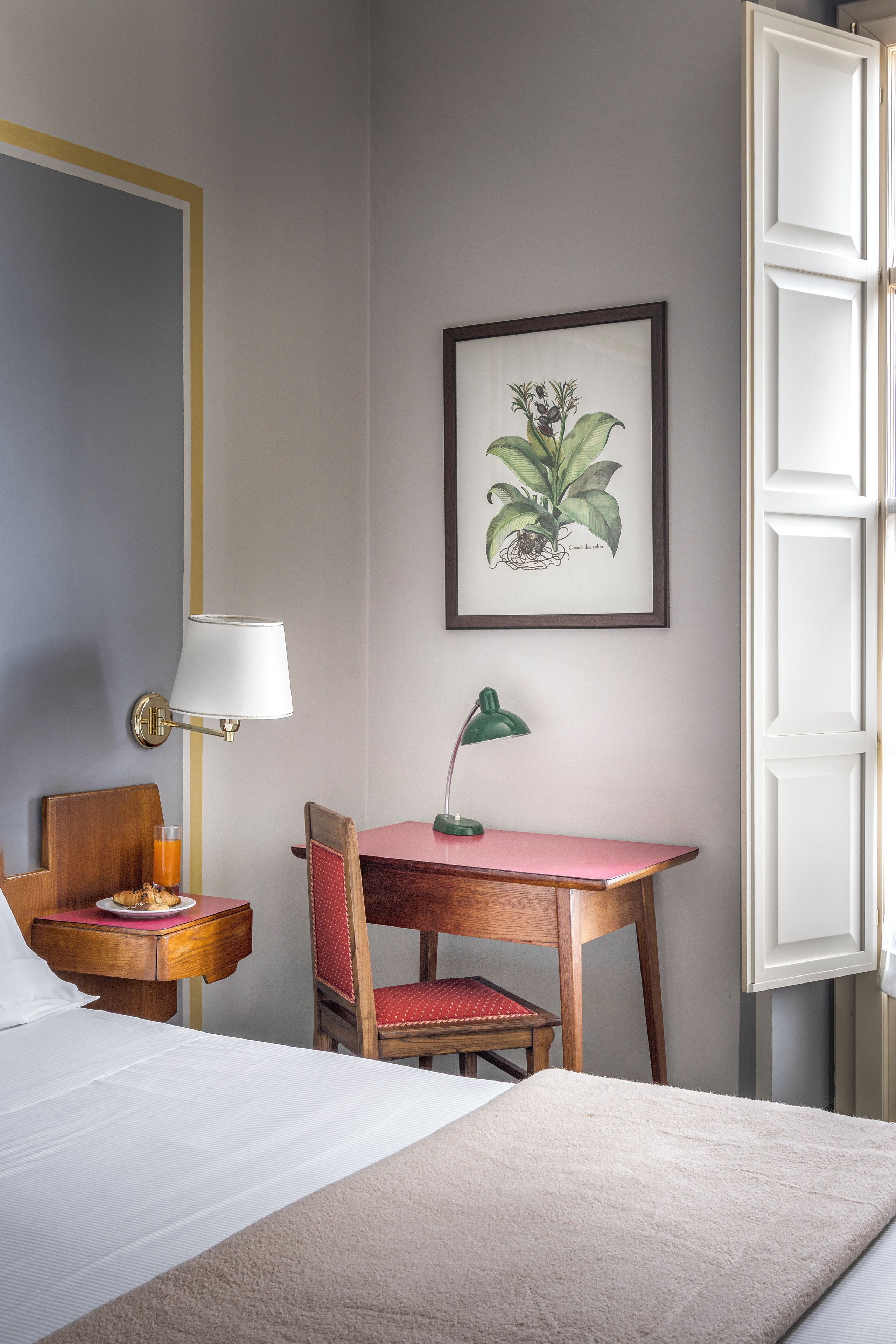 Quarto de hotel com cama, escrivaninha, cadeira, luminária e ilustração botânica emoldurada na parede.