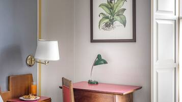 Quarto de hotel com cama, escrivaninha, cadeira, luminária e ilustração botânica emoldurada na parede.