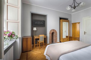 Quarto de hotel com cama, escrivaninha, poltrona, espelho, lareira e placa fixada na parede.