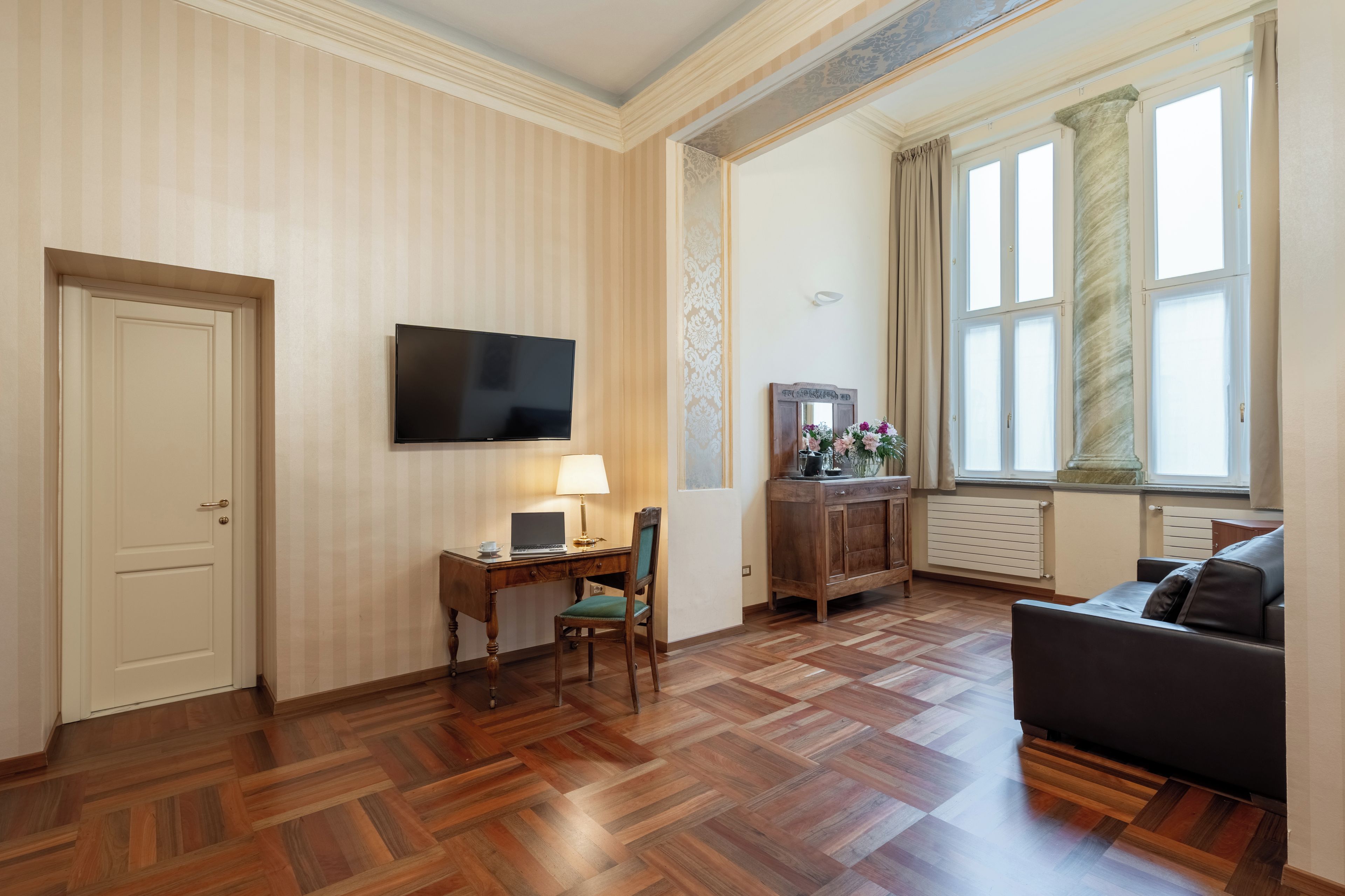 Quarto de hotel com chão em assoalho de madeira, televisão de tela plana na parede, escrivaninha com laptop e luminária, armário de madeira com espelho e sofá.