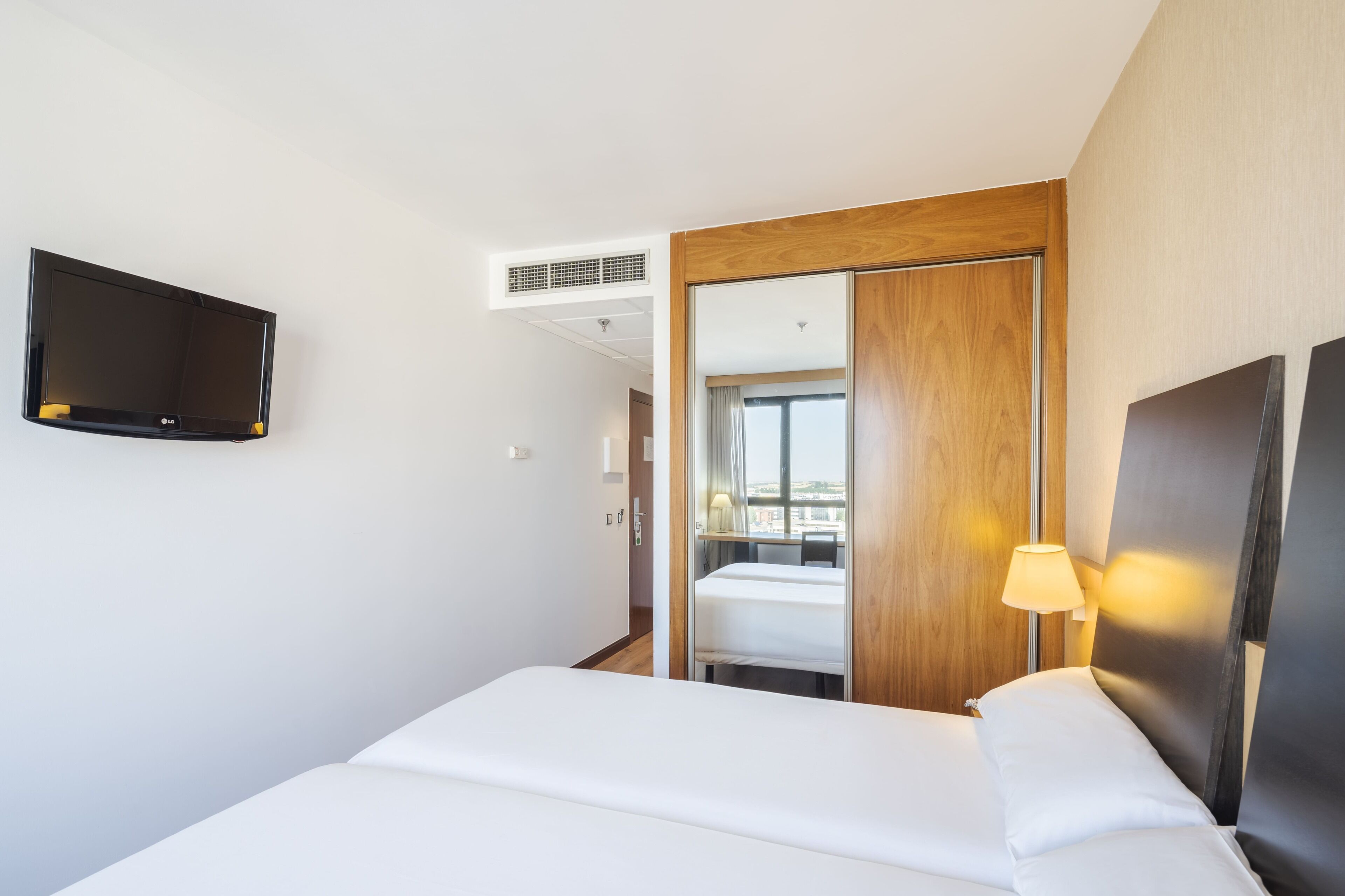 Quarto de hotel com uma cama, televisão fixada na parede, porta de madeira e janela com vista.