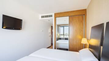 Quarto de hotel com uma cama, televisão fixada na parede, porta de madeira e janela com vista.