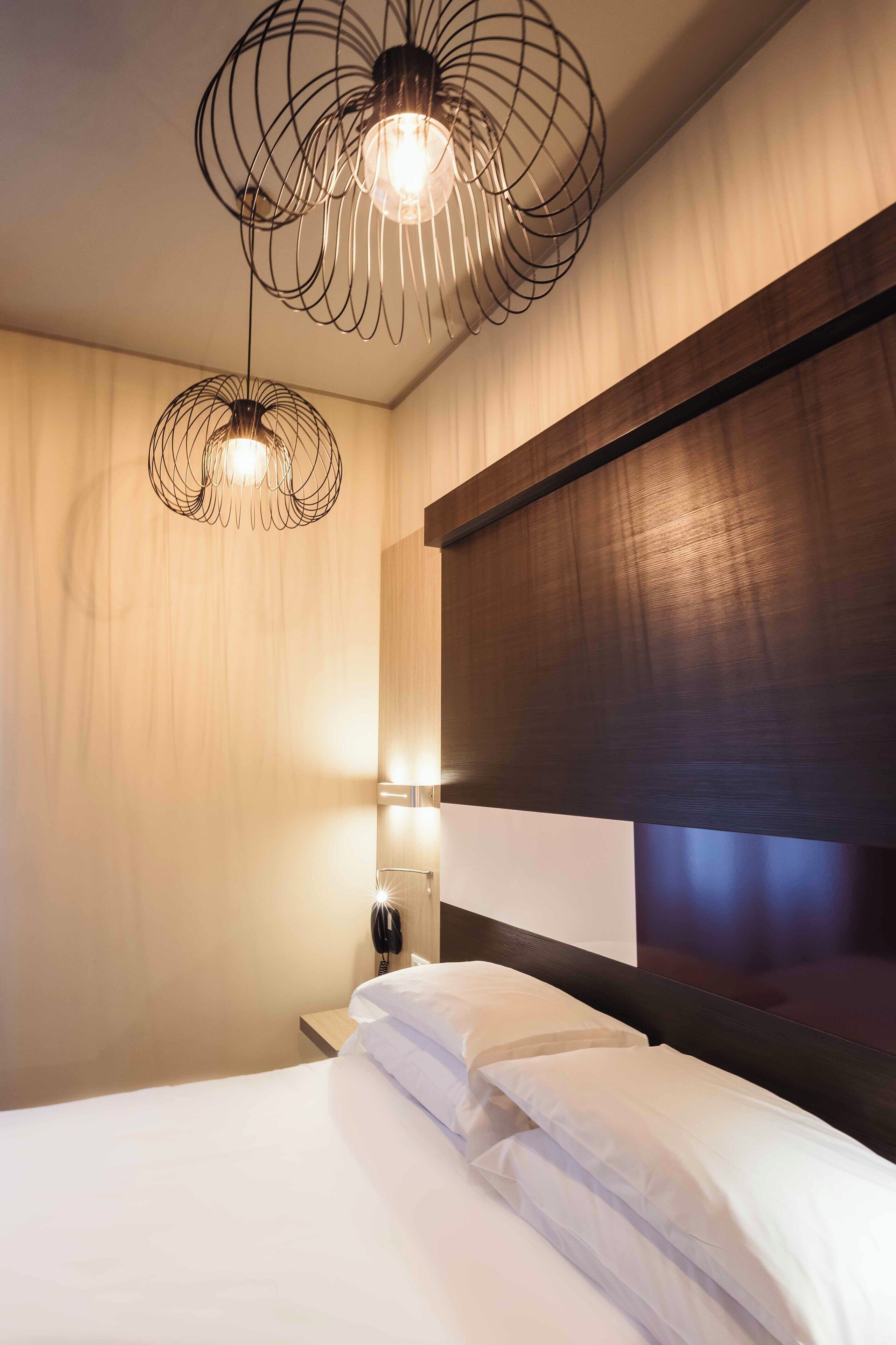 Quarto de hotel com cama, duas luminárias pendentes, telefone fixado na parede e cabeceira de madeira.