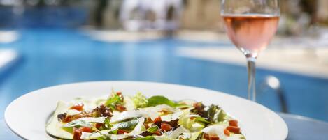 Uma salada num prato, com um copo de vinho rosé, sobre uma mesa perto de uma piscina.
