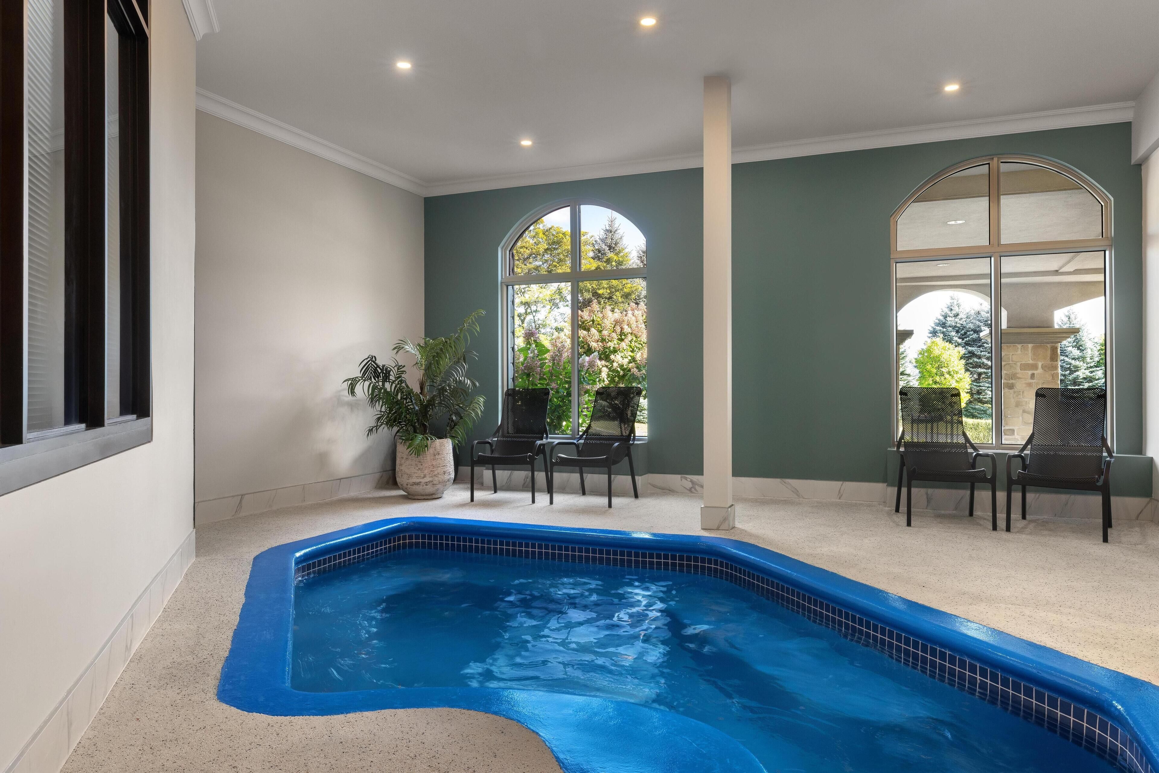 Piscina interior com borda azulejada azul, rodeada por piso bege e parede verde. Há duas cadeiras pretas e uma planta em vaso perto de uma grande janela em arco.