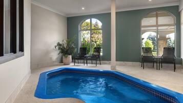 Piscina interior com borda azulejada azul, rodeada por piso bege e parede verde. Há duas cadeiras pretas e uma planta em vaso perto de uma grande janela em arco.