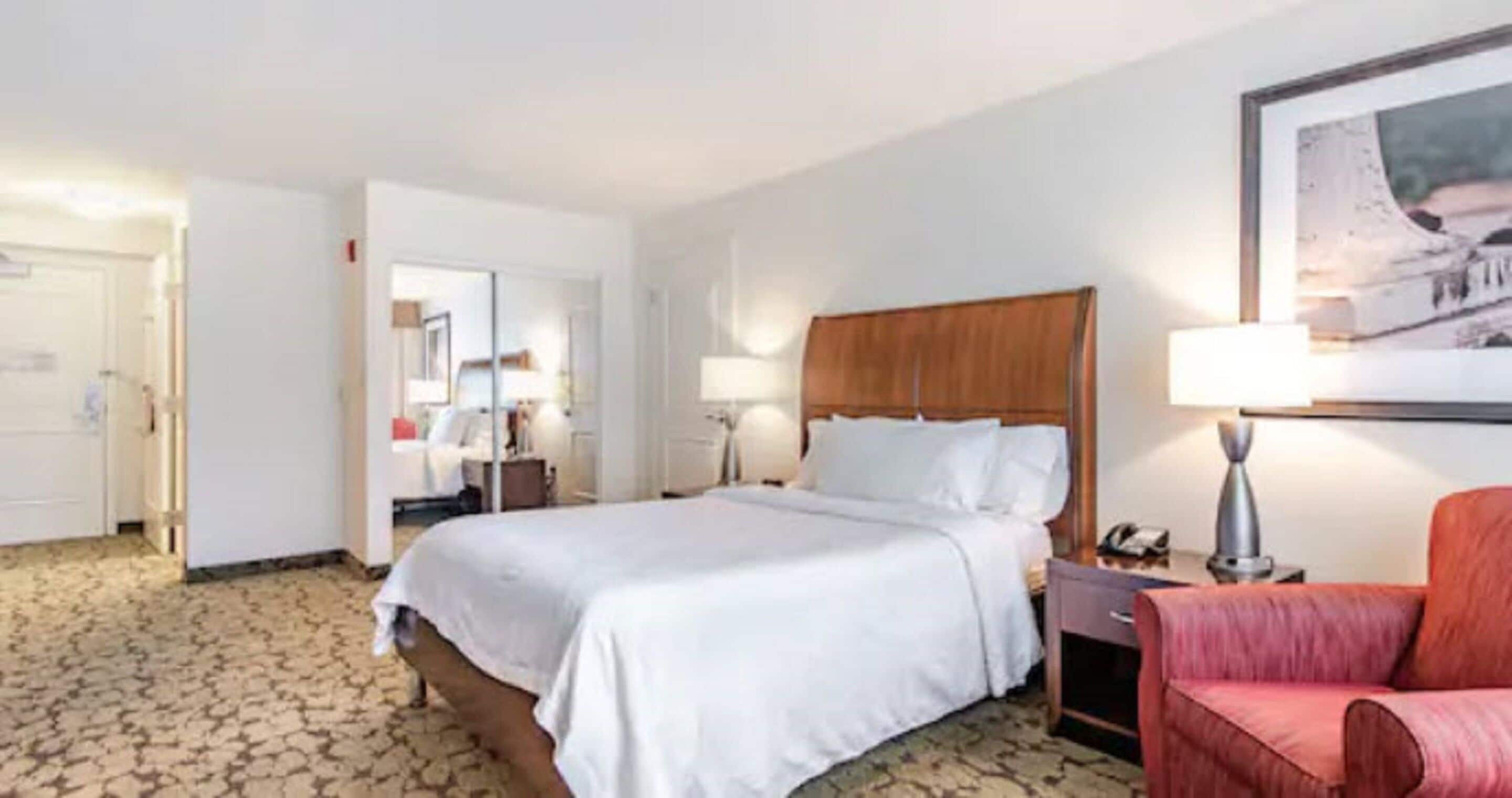 Quarto de hotel com cama, mesa de cabeceira, abajur, cadeira e um quadro na parede.