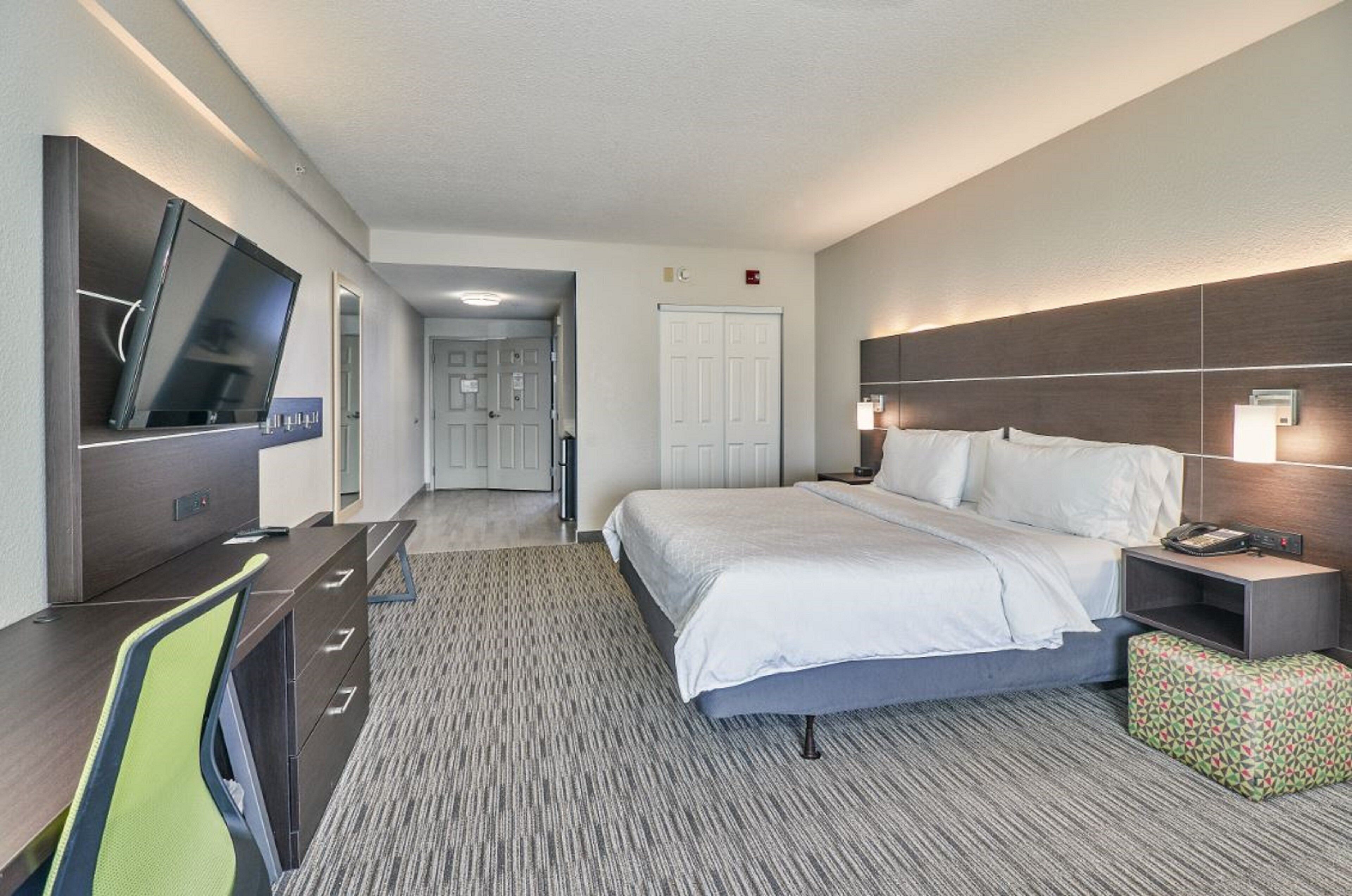 Quarto de hotel com cama, mesa de cabeceira, luminária, escrivaninha com cadeira, televisão e porta para outro cômodo.