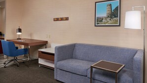 Quarto de hotel com sofá azul, uma mesa com cadeira, luminária e quadro pendurado na parede.