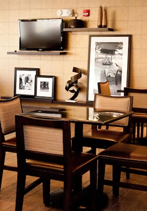 Uma sala de jantar com mesa de vidro, cadeiras de madeira, televisão fixada numa prateleira e quadros nas paredes.