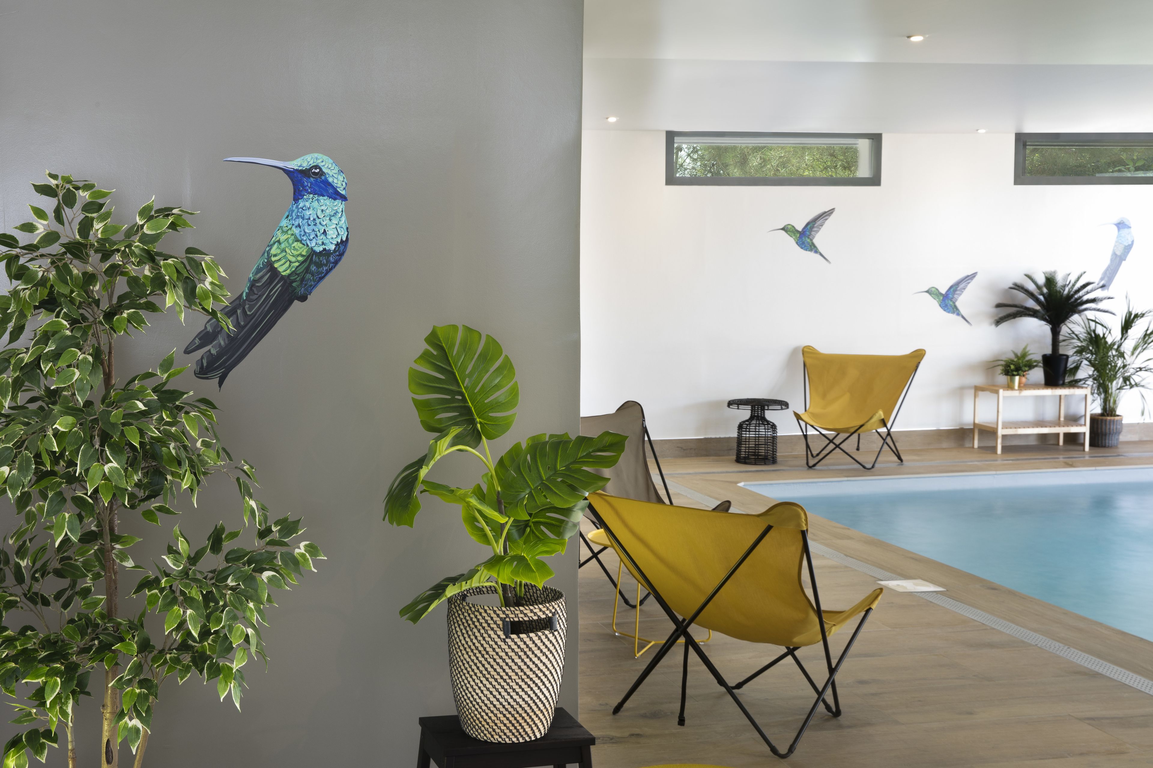 Uma área interna de piscina moderna com um grande mural na parede retratando pássaros, uma planta em vaso e cadeiras amarelas.
