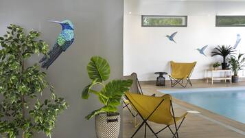 Uma área interna de piscina moderna com um grande mural na parede retratando pássaros, uma planta em vaso e cadeiras amarelas.