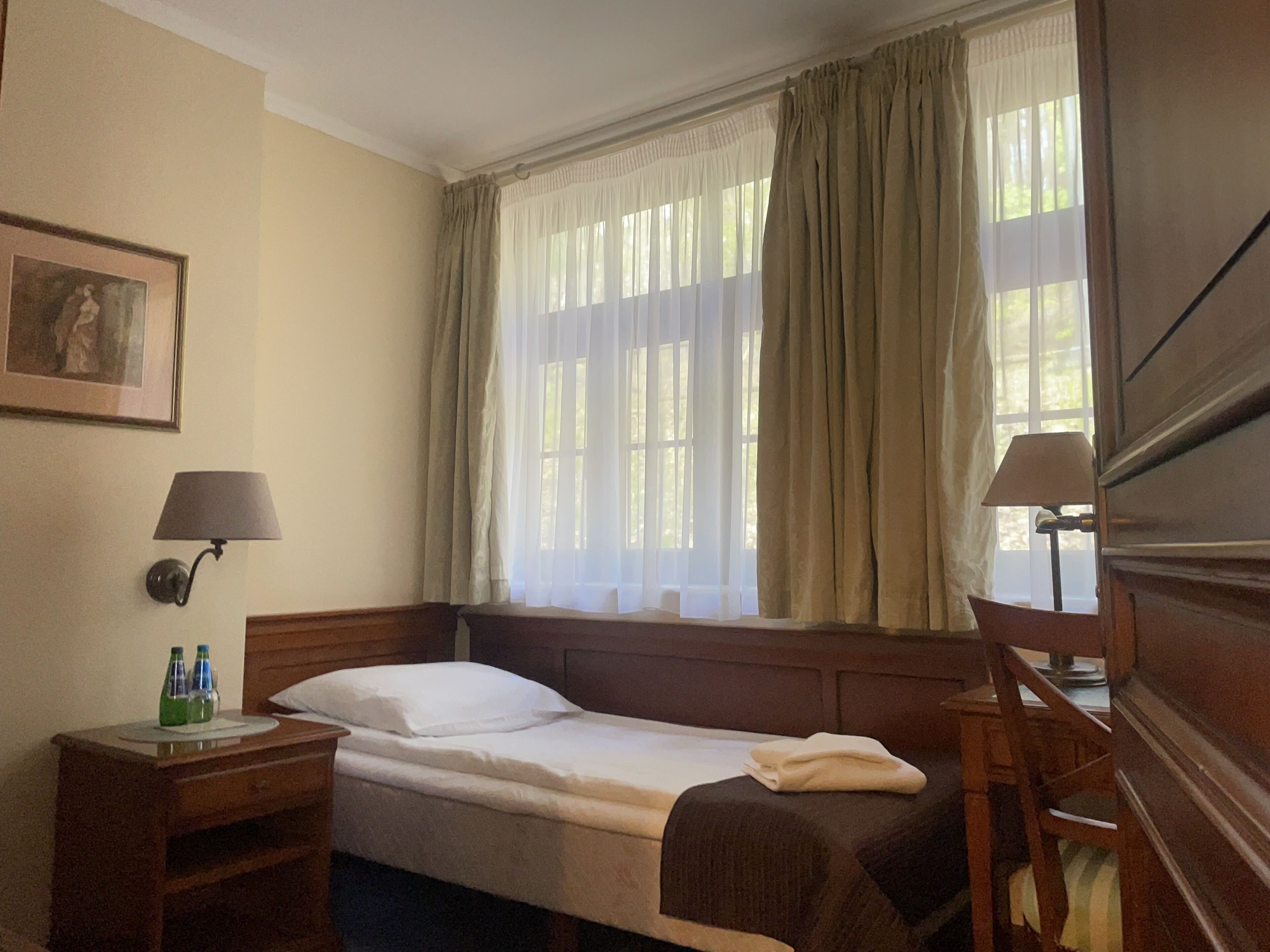 Quarto de hotel com uma cama de solteiro, mesa de cabeceira com abajur, uma cadeira, janela com cortinas e um quadro na parede.