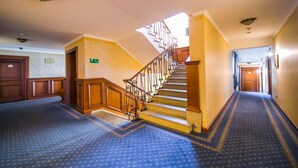 Um corredor de hotel com carpete azul, escada de madeira com guarda-corpo de ferro forjado e um claraboia acima.