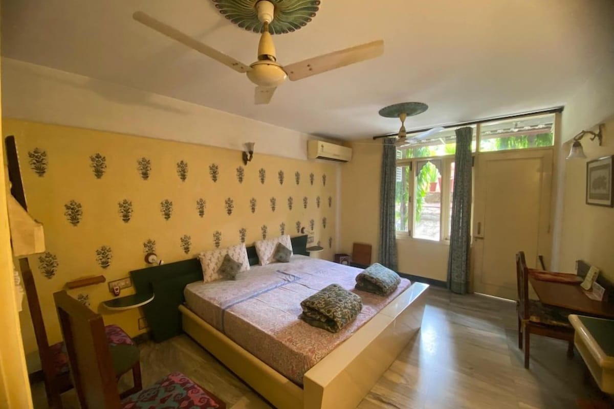 Quarto com cama, escrivaninha, cadeira, ventoinha de teto e janela.