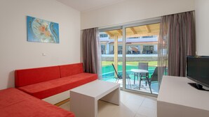 Quarto moderno com sofá vermelho, mesa de centro branca e TV. Possui porta de vidro que dá para uma área externa com piscina e cadeiras.