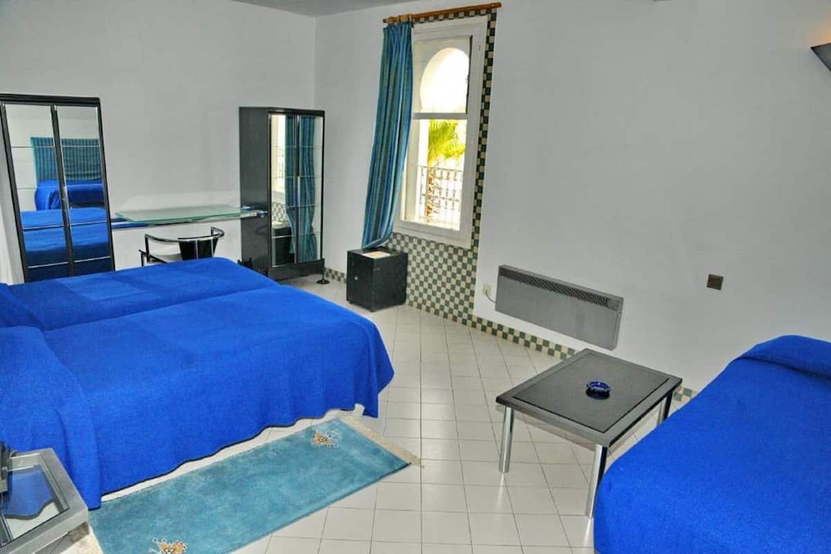 Quarto de hotel com duas camas, uma mesa pequena, uma cadeira, um espelho e uma janela com cortinas.