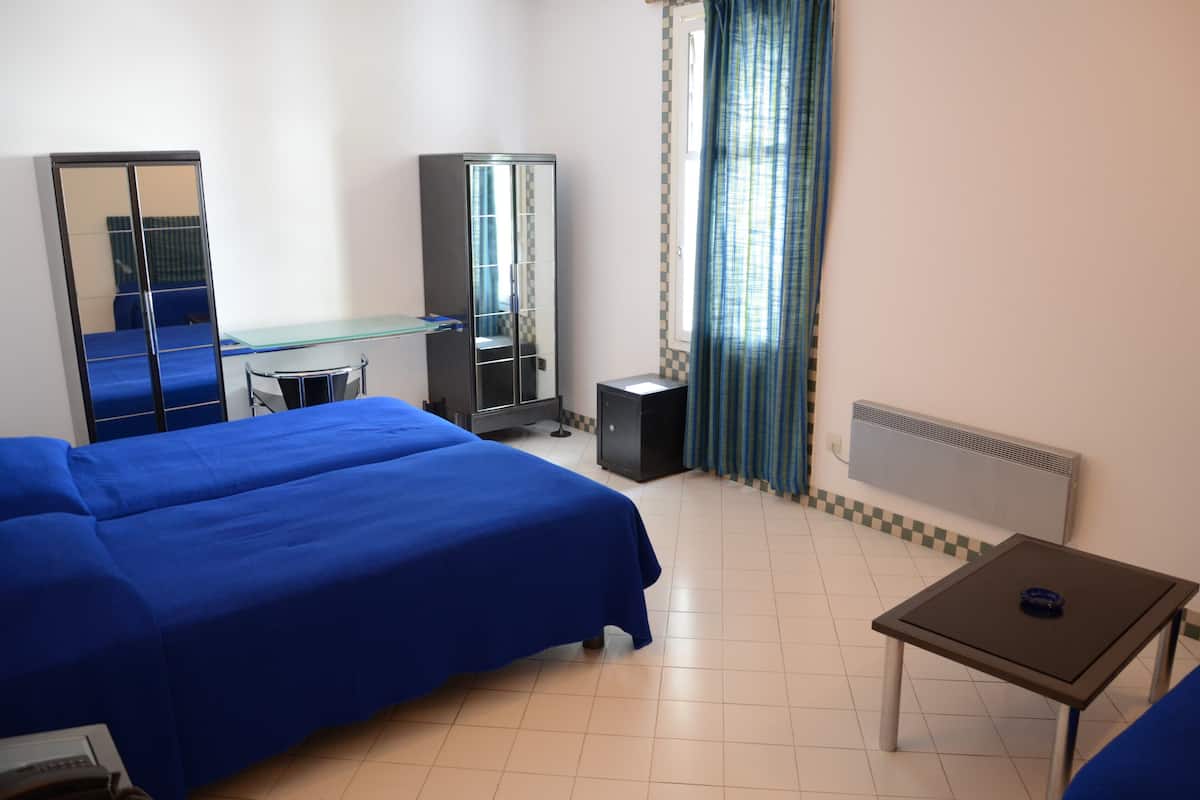 Quarto de hotel com duas camas, uma escrivaninha, uma cadeira, um espelho, um radiador e uma mesinha.