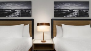 Quarto de hotel com duas camas, uma mesinha de cabeceira com abajur e duas imagens emolduradas na parede.