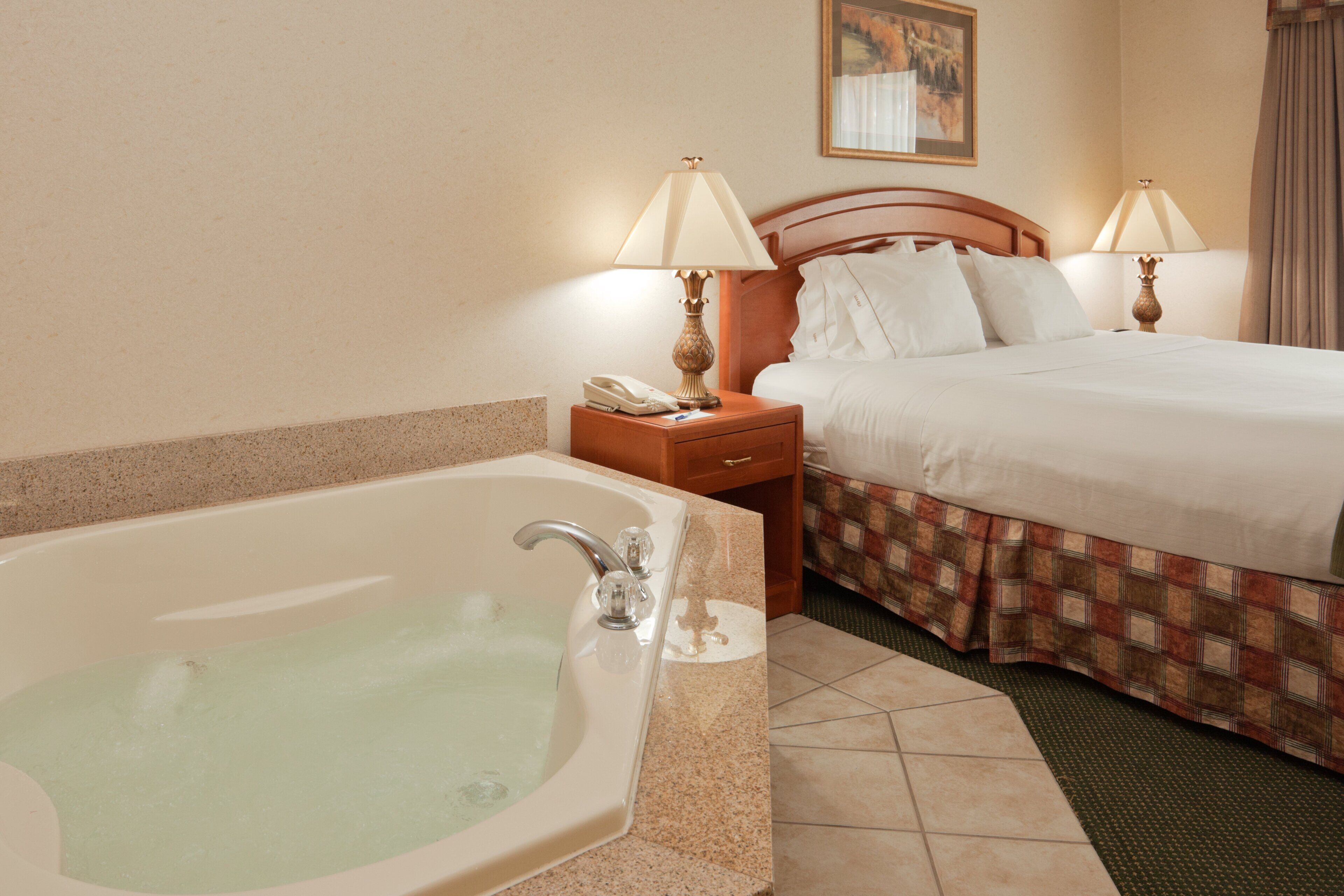 Quarto de hotel com uma banheira grande, cama com roupa de cama branca, duas luminárias de cabeceira e um quadro na parede.
