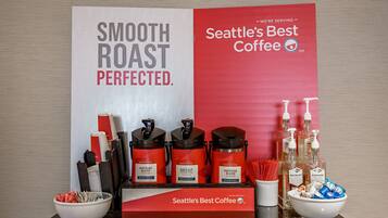 Um espaço com estação de café, dispensadores de café identificados, um recipiente com lanches e uma placa com a inscrição 'Seattle's Best Coffee'.