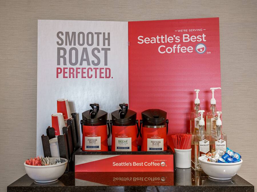 Um espaço com estação de café, dispensadores de café identificados, um recipiente com lanches e uma placa com a inscrição 'Seattle's Best Coffee'.