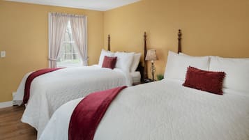 Quarto de hotel com duas camas, uma janela com cortinas, mesa de cabeceira com abajur e chão de madeira.